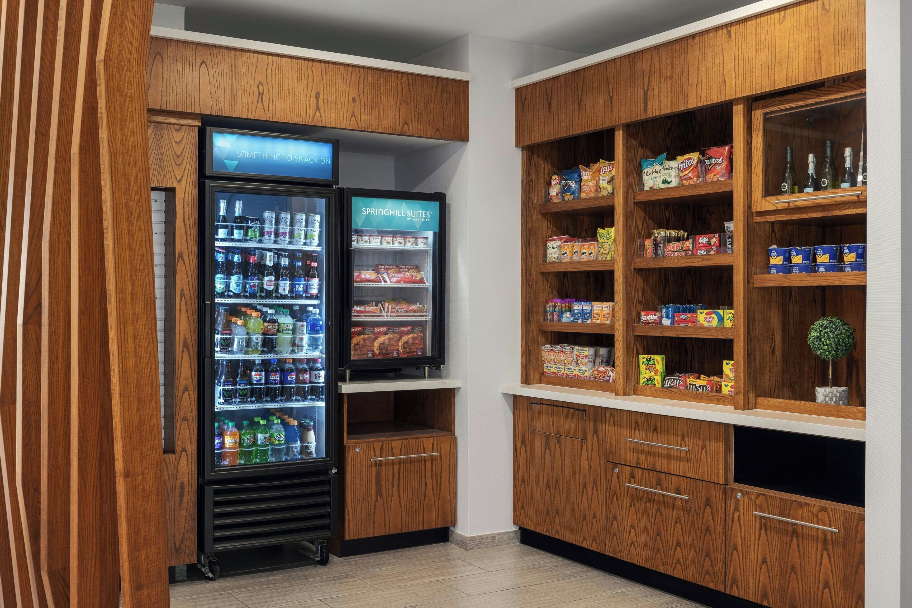 Um canto de uma loja com armários de madeira, uma vitrine refrigerada e prateleiras repletas de lanches e bebidas.