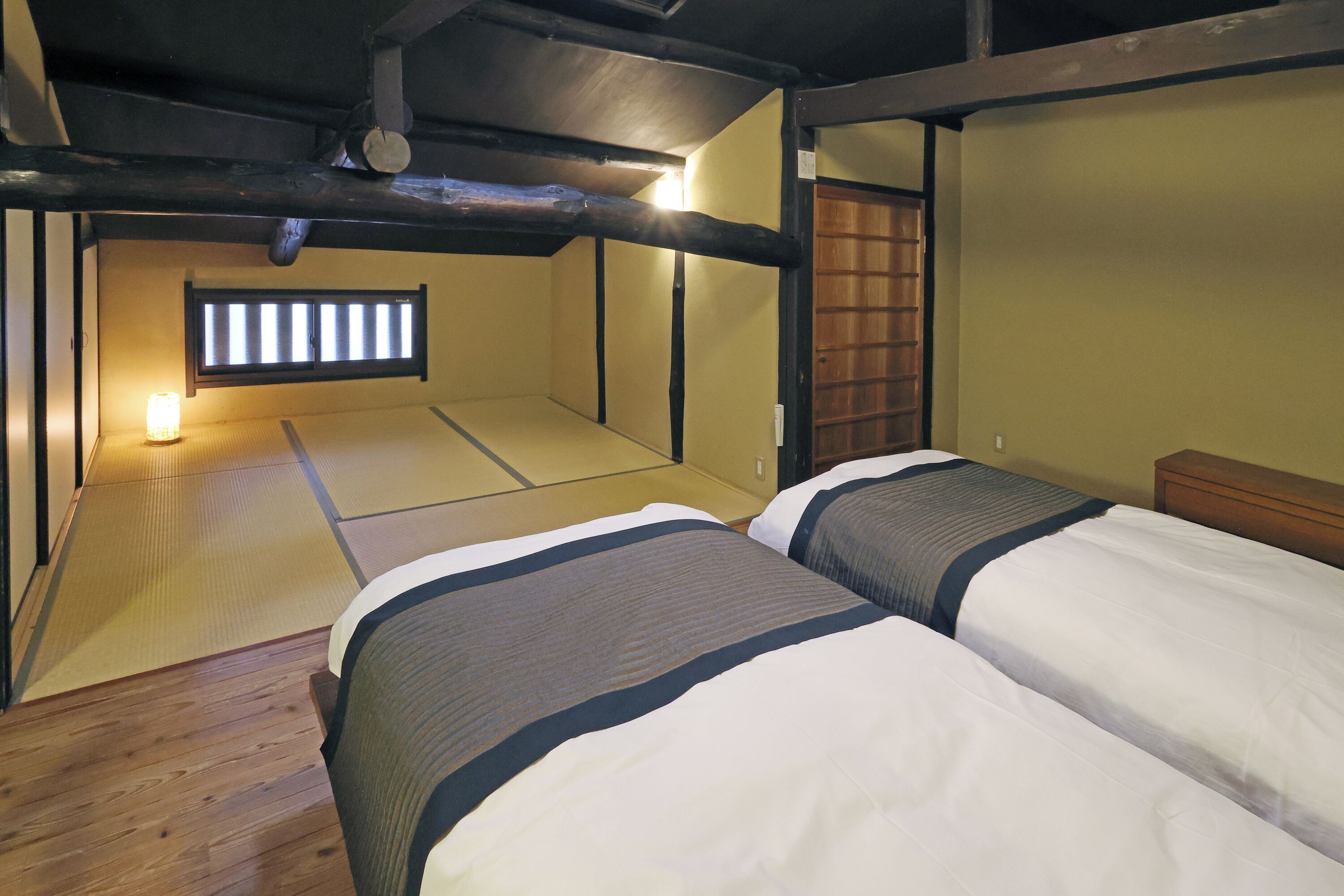 Quarto japonês tradicional com tatames, duas camas com roupa de cama escura e branca, teto de madeira e porta deslizante.