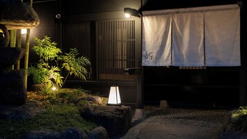 Vista noturna de um edifício tradicional japonês com jardim de bambu, um lanterna e uma cortina branca.