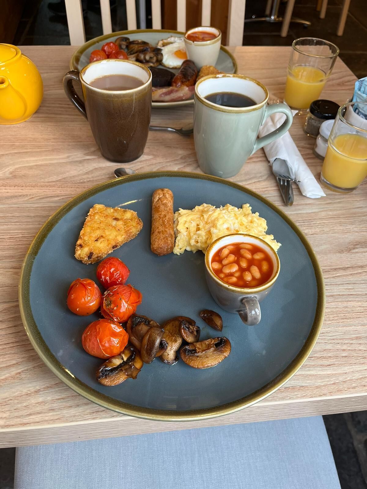 Mesa de café da manhã com prato de ovos, feijão, cogumelos e tomate, acompanhado de café e suco.
