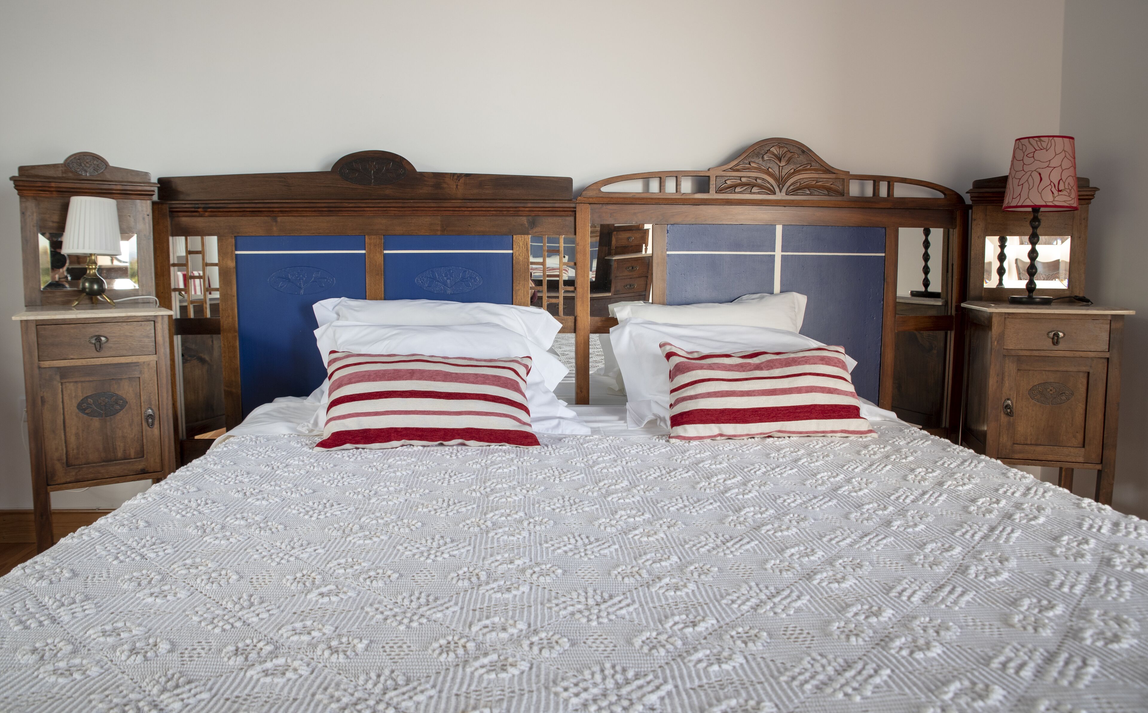Cama de casal com colcha branca texturizada e duas almofadas, uma listrada e outra branca lisa, em quarto com cabeceira e mesas de cabeceira de madeira.