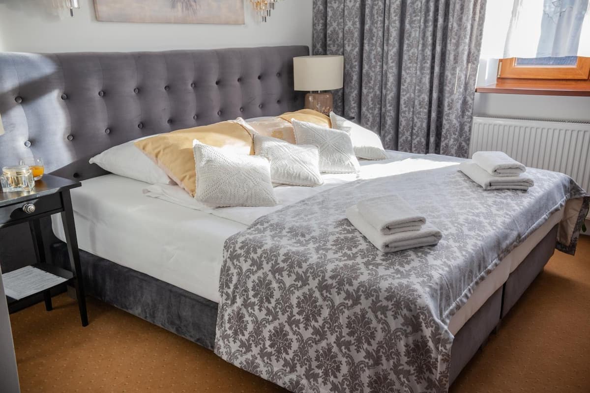 Uma cama bem arrumada com cabeceira cinza, roupa de cama branca e estampada, e uma mesa de cabeceira com abajur.
