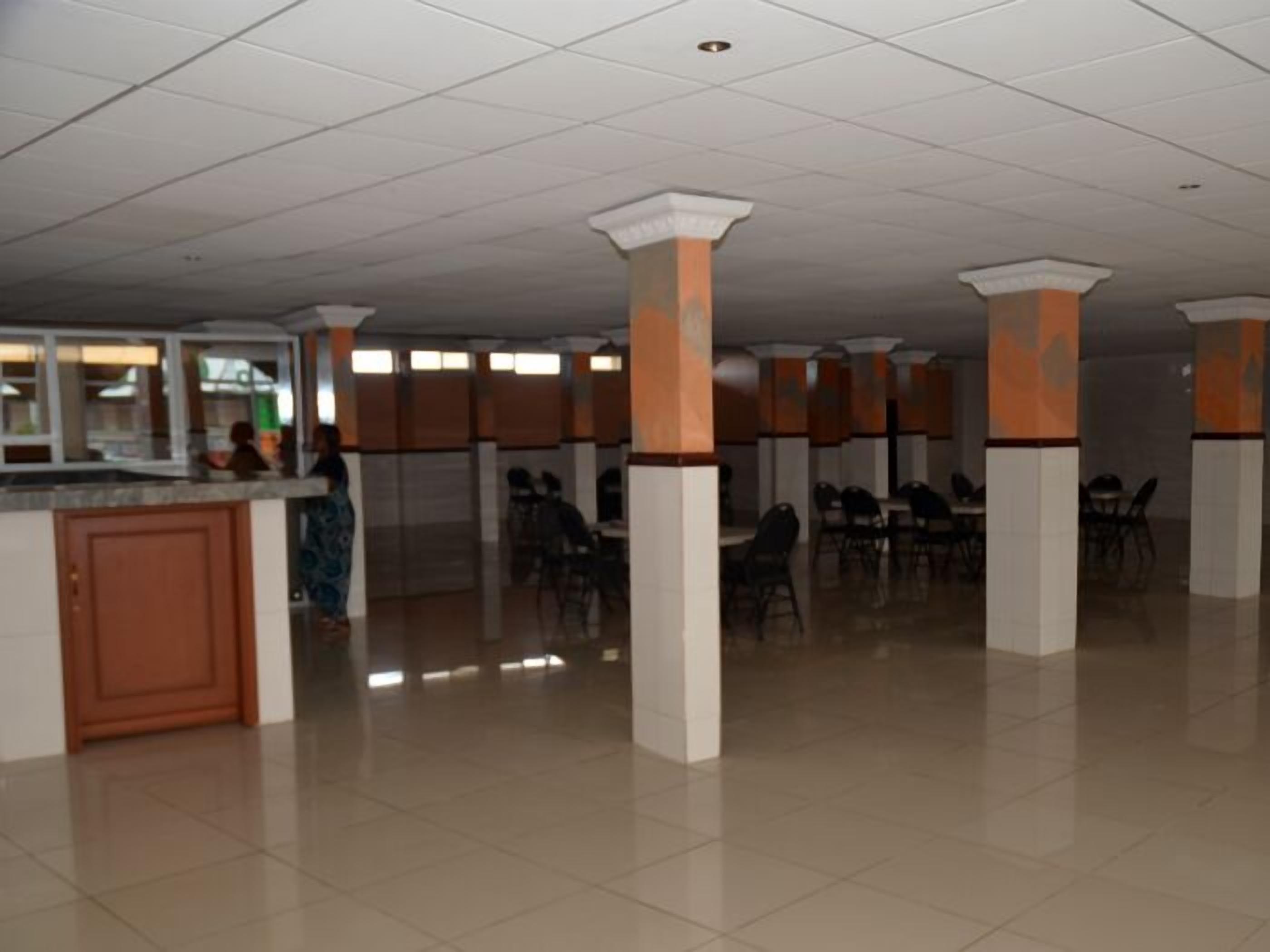 Um espaço amplo e interno, com piso ladrilhado, várias mesas e cadeiras, e um balcão com armário de madeira.