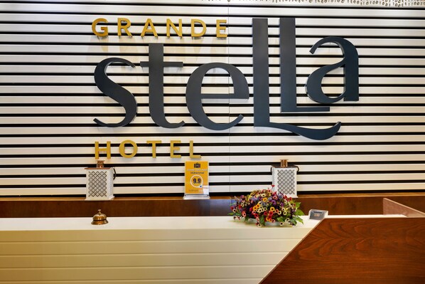 Uma receção de hotel com uma placa a indicar "GRANDE stella HOTEL" e um preço de 9,9$ por noite.