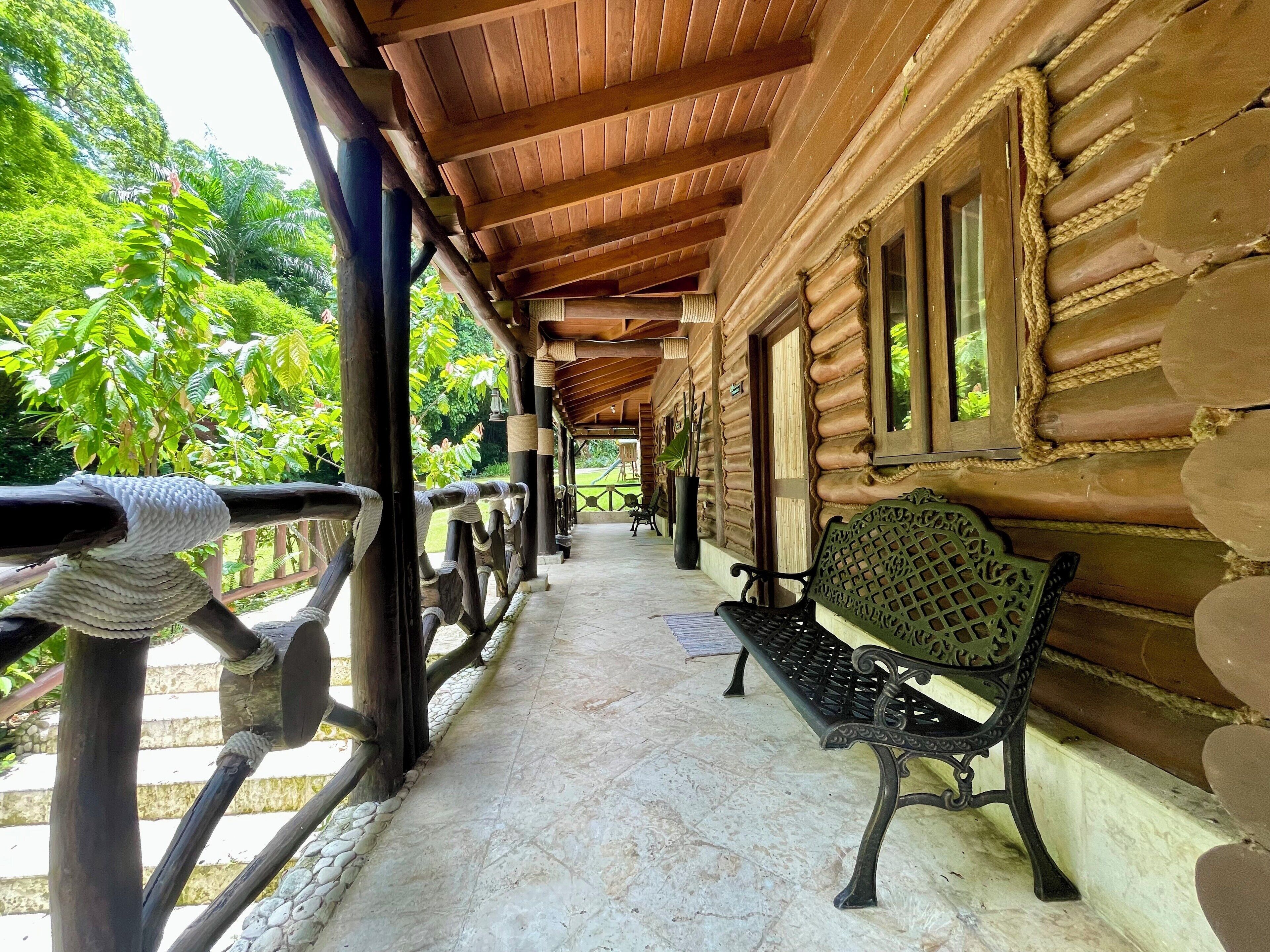 Uma passarela de madeira coberta com um banco e guarda-corpo de corda, que conduz a uma cabana de troncos com grandes janelas.