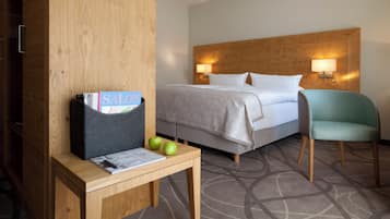 Quarto de hotel com cabeceira de madeira, duas luminárias de apoio, cama com lençóis brancos, uma mesinha com revista e maçãs, e uma cadeira azul-turquesa.