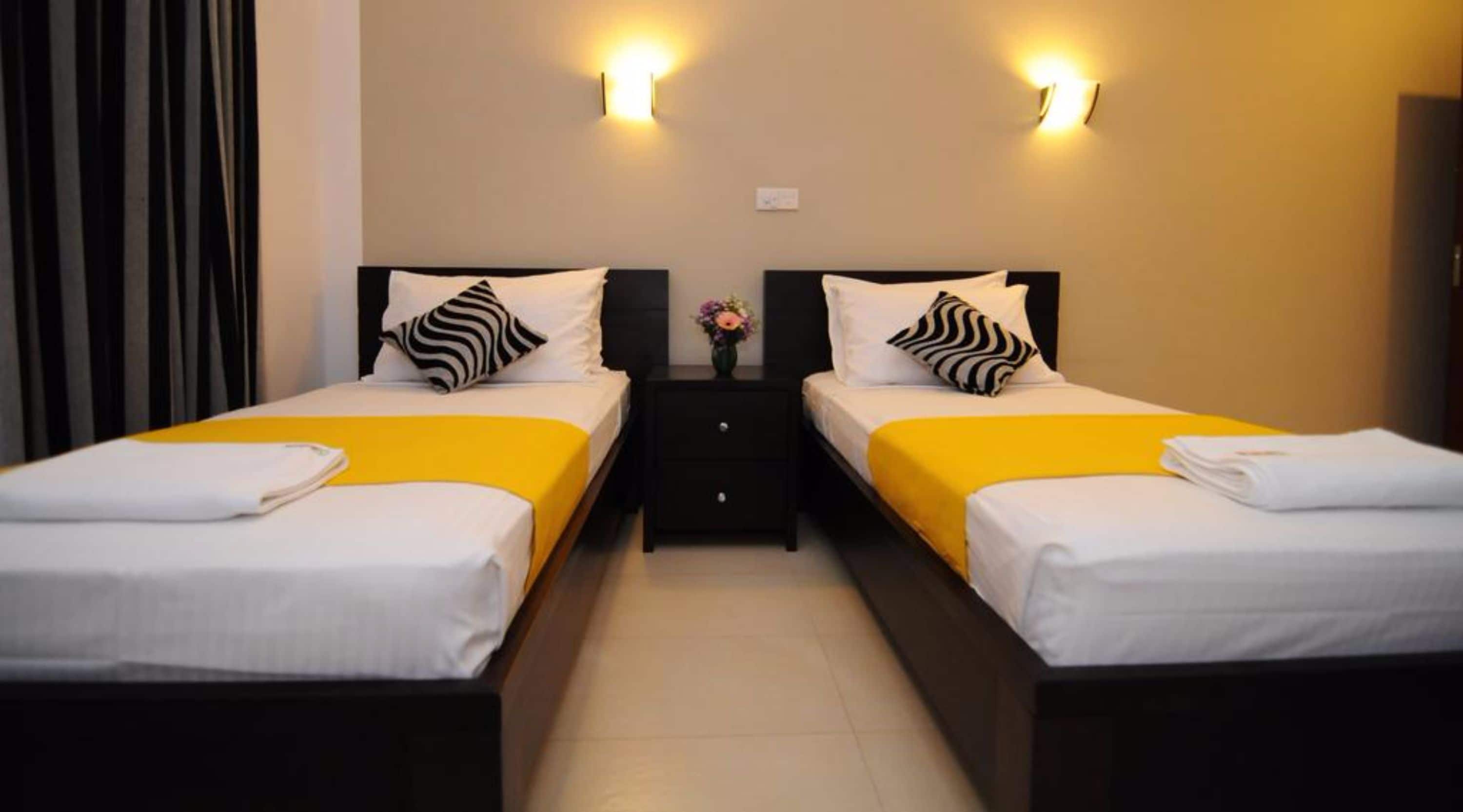 Quarto de hotel com duas camas de solteiro, cada uma com colcha branca e detalhe amarelo. Há duas mesas de cabeceira com superfície preta, uma entre as camas e outra à direita. Cada cama tem um travesseiro listrado em preto e branco.