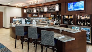Uma área de bar com balcão curvo, três banquetas, uma televisão na parede e diversas garrafas e copos nas prateleiras.