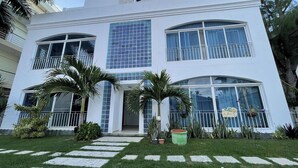 Uma casa branca de dois andares com grandes janelas, varanda frontal e gramado bem cuidado.