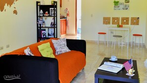 Uma sala de estar com um sofá, uma mesa de centro e uma pequena mesa com um laptop e uma bandeira americana. Há uma estante com diversos objetos e um mapa-múndi na parede.