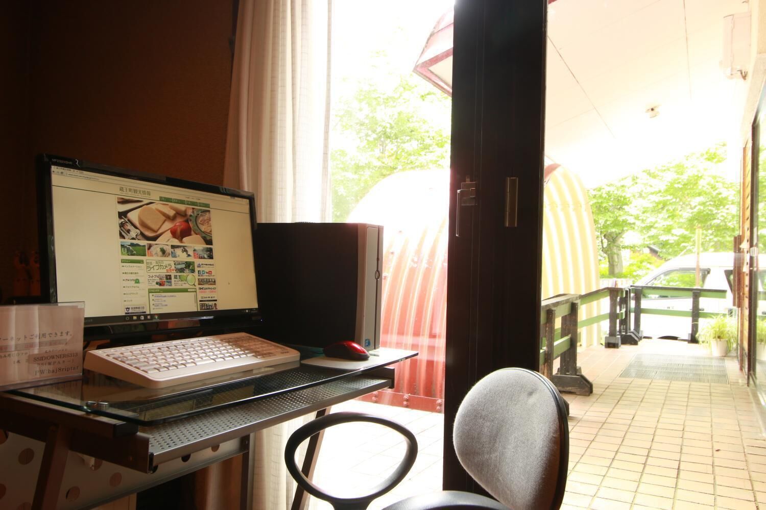 Quarto com escrivaninha, computador e cadeira. Há uma janela com cortinas, uma porta e vista para o exterior.