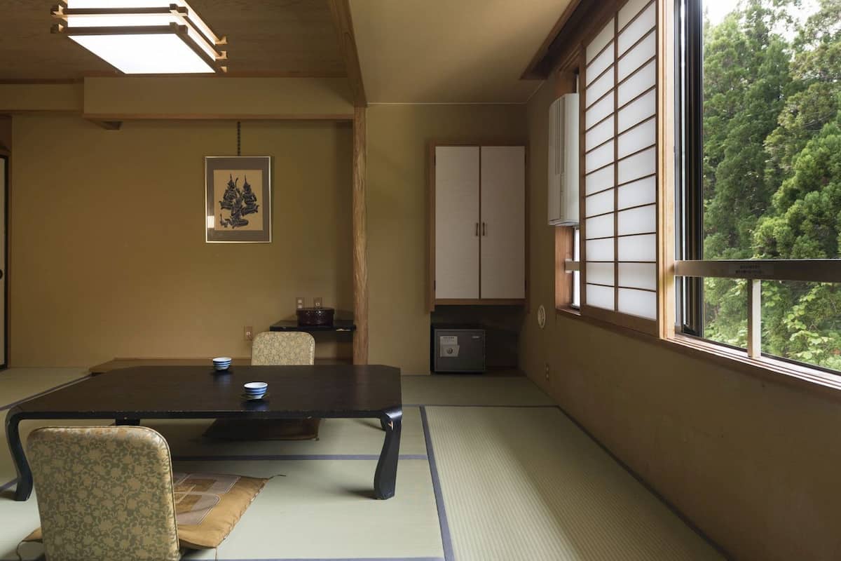 Um espaço com tatames, uma mesa baixa, cadeiras, um armário e uma janela grande com vista para árvores.