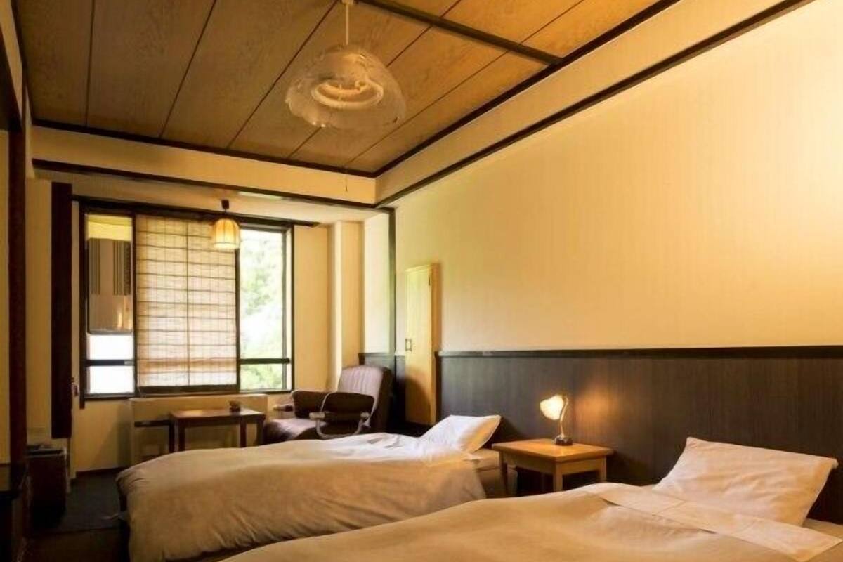 Quarto de hotel com duas camas, uma cadeira, uma escrivaninha e uma janela com persianas.