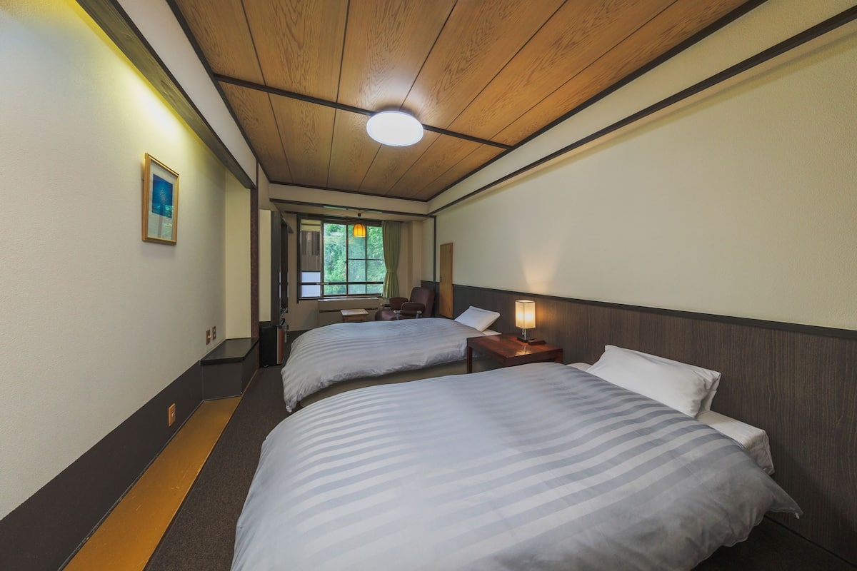 Quarto de hotel com duas camas, teto de madeira, janela com vista e uma pequena mesa com abajur.