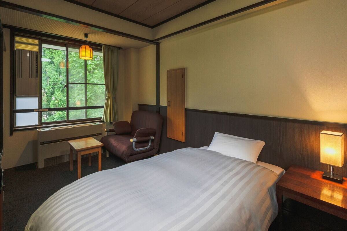 Quarto de hotel com cama, cadeira, escrivaninha e janela com vista para árvores.