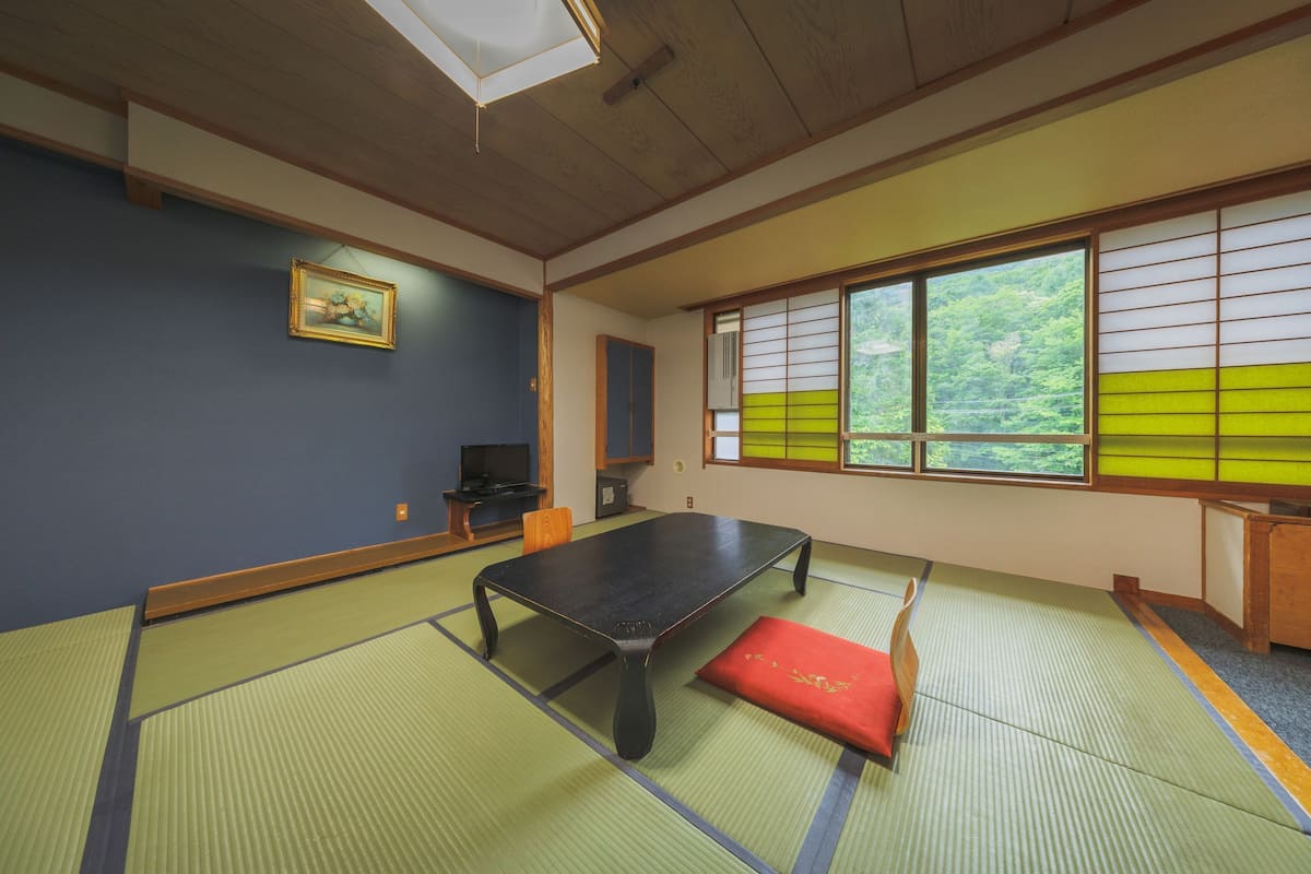 Um espaço com tatames, uma mesa baixa, um assento de almofada, uma televisão e uma janela com divisórias shoji.