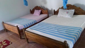 Duas camas de madeira com roupa de cama listrada em azul, cabeceira de madeira e um tapete decorativo vermelho sobre um chão ladrilhado.