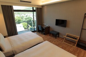 Quarto de hotel com duas camas, uma escrivaninha, uma cadeira, televisão e varanda com vista para áreas verdes e edifícios.
