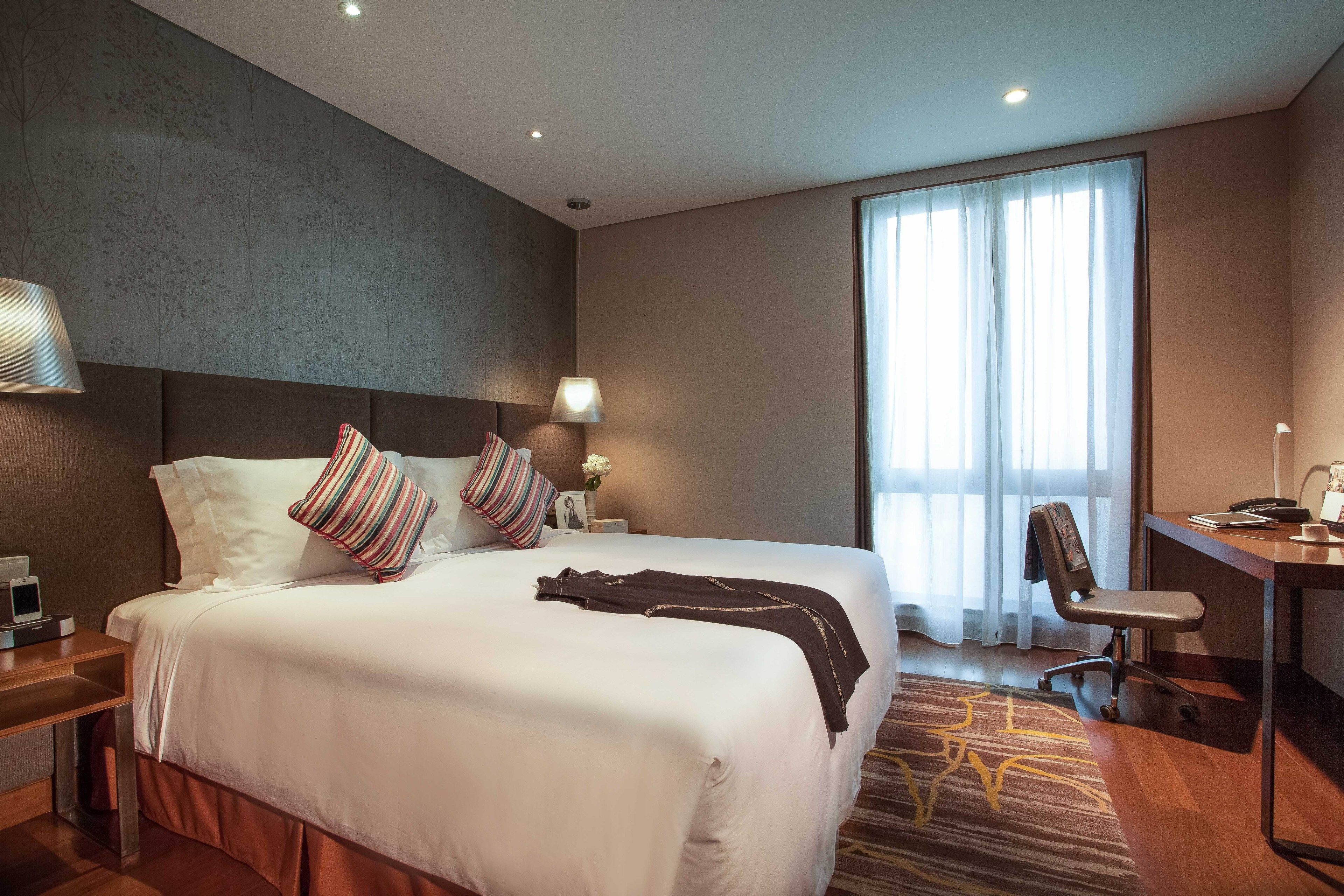 Quarto de hotel com uma cama grande, uma mesa de trabalho e uma cadeira. Há uma janela com cortinas, uma luminária e uma mesa de cabeceira com um telefone e um vaso com flores.