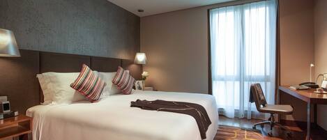 Quarto de hotel com uma cama grande, uma mesa de trabalho e uma cadeira. Há uma janela com cortinas, uma luminária e uma mesa de cabeceira com um telefone e um vaso com flores.