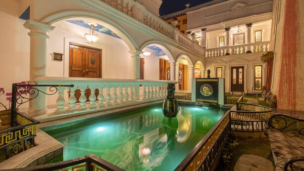 Uma área de piscina com uma fonte, um espaço de estar com sofá e cadeiras, e uma varanda com guarda-corpo.