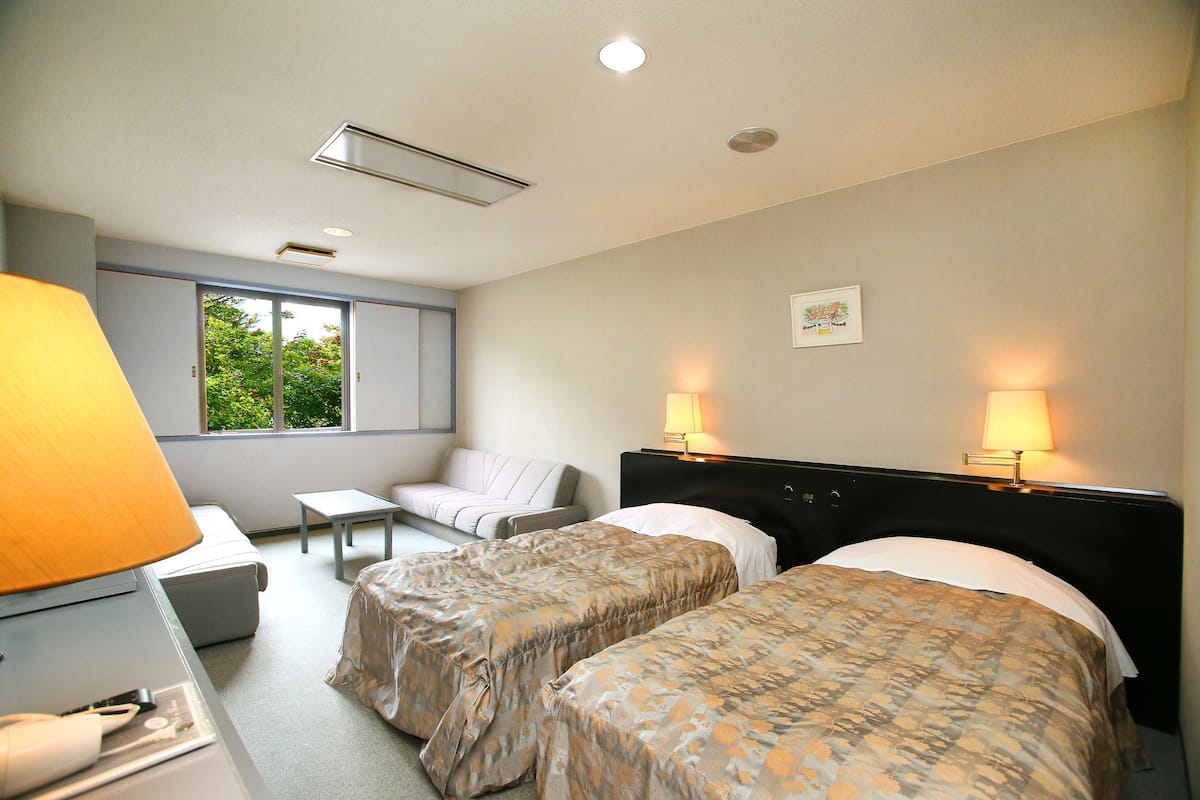 Quarto de hotel com duas camas, um sofá, uma mesa pequena e uma luminária.