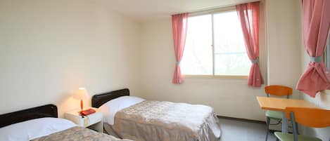 Quarto de hotel com duas camas, uma escrivaninha, uma cadeira e uma janela com cortinas.