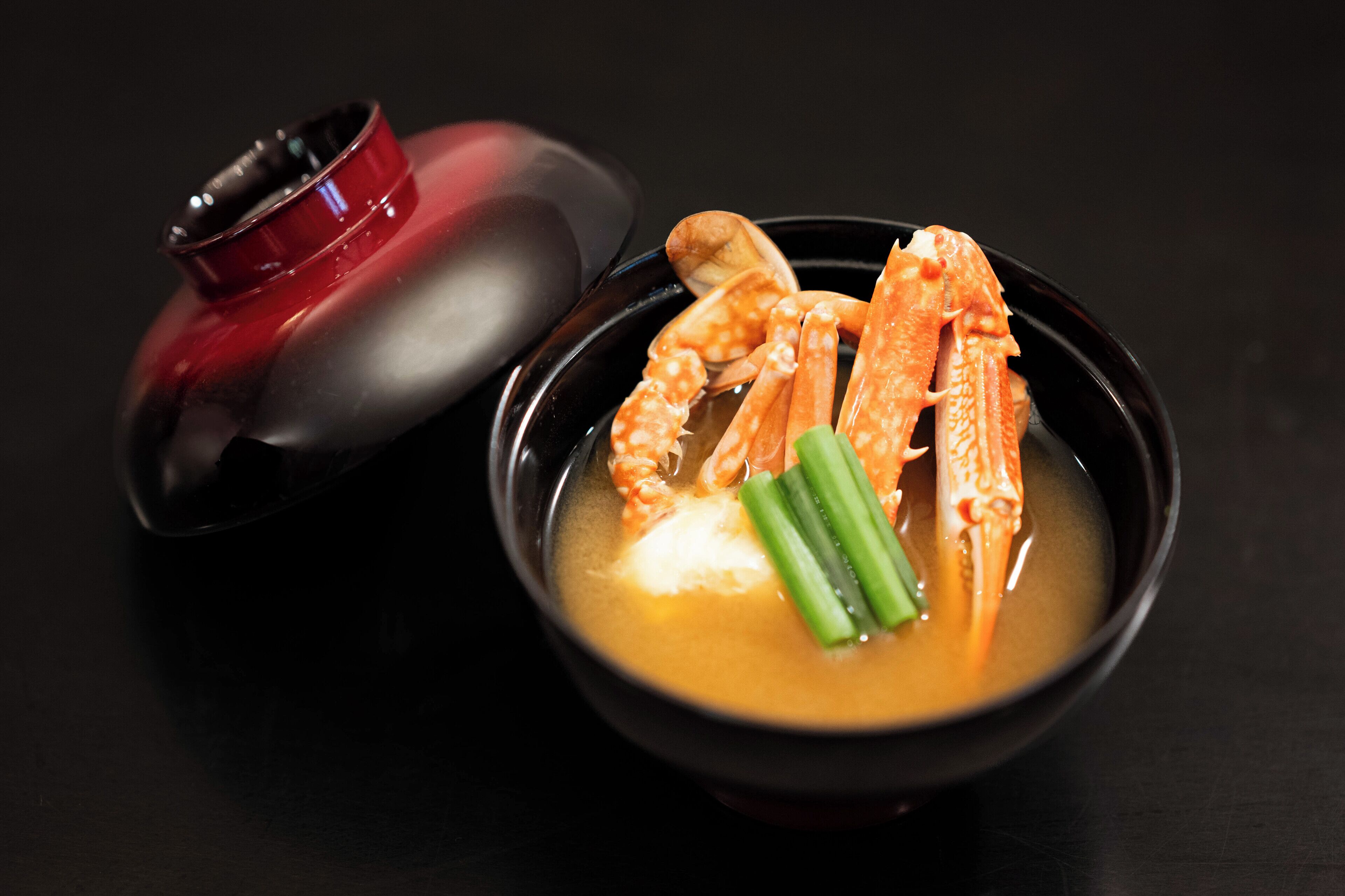 Uma tigela de sopa com patas de caranguejo e cebolinha, acompanhada por um recipiente vermelho e preto.