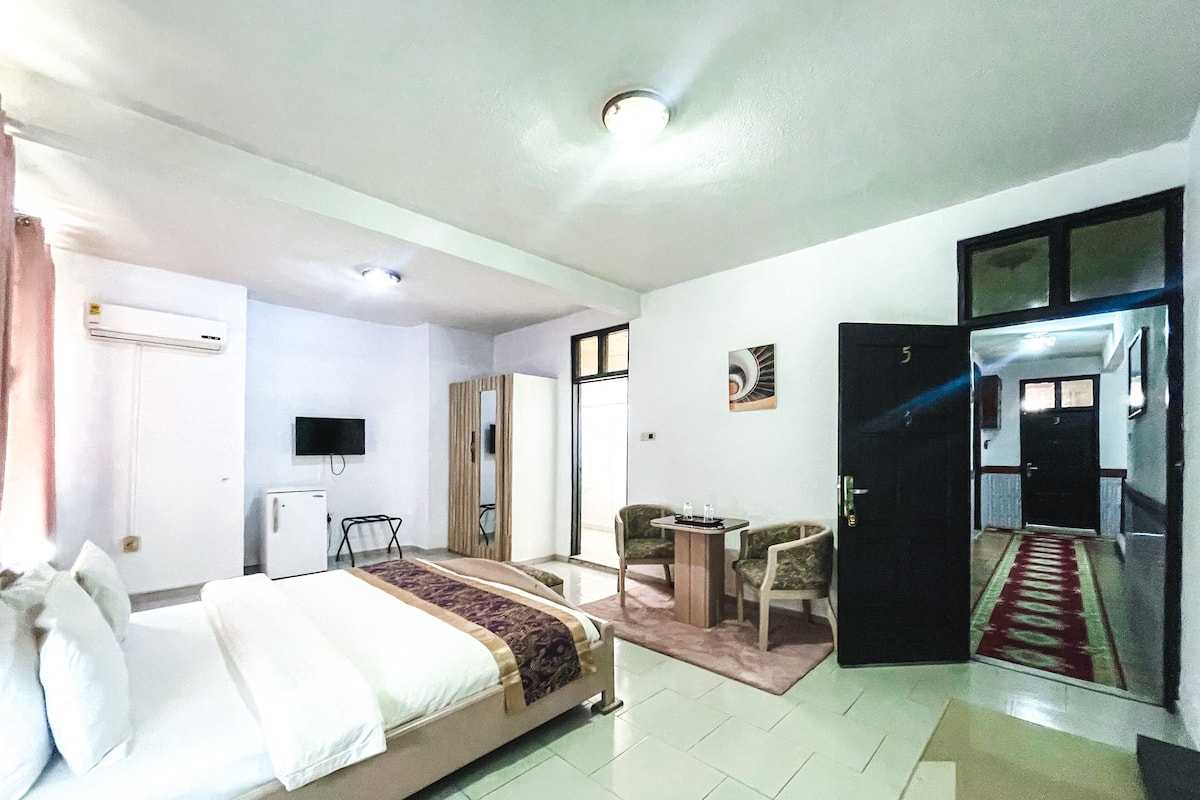 Quarto de hotel com cama, escrivaninha, duas cadeiras, espelho, televisão e porta que dá para um corredor.