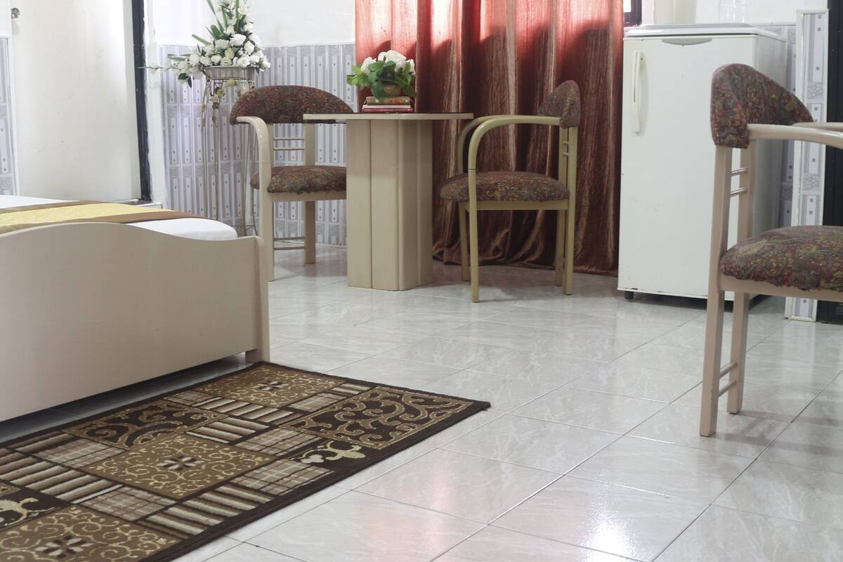 Quarto com cama, escrivaninha, duas cadeiras, frigorífico e janela com cortinas.