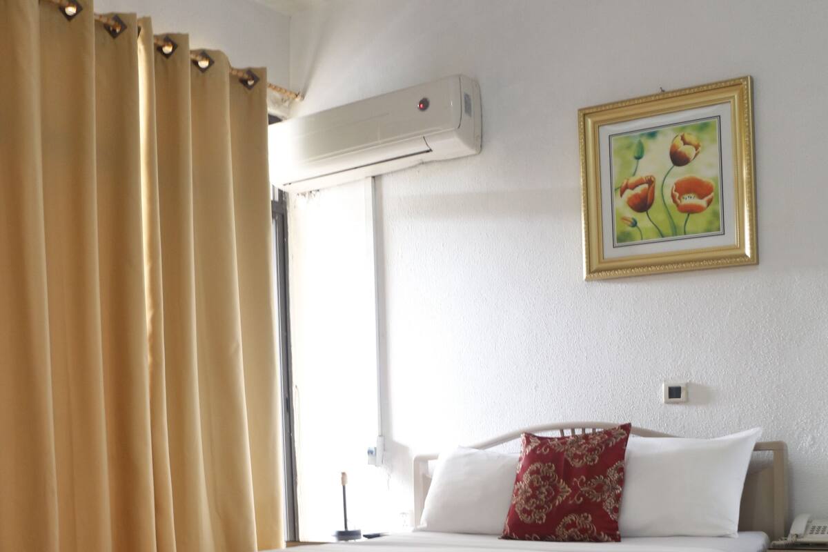 Quarto de hotel com cama, mesa de cabeceira, cortinas na janela, ar condicionado fixado na parede e um quadro emoldurado.
