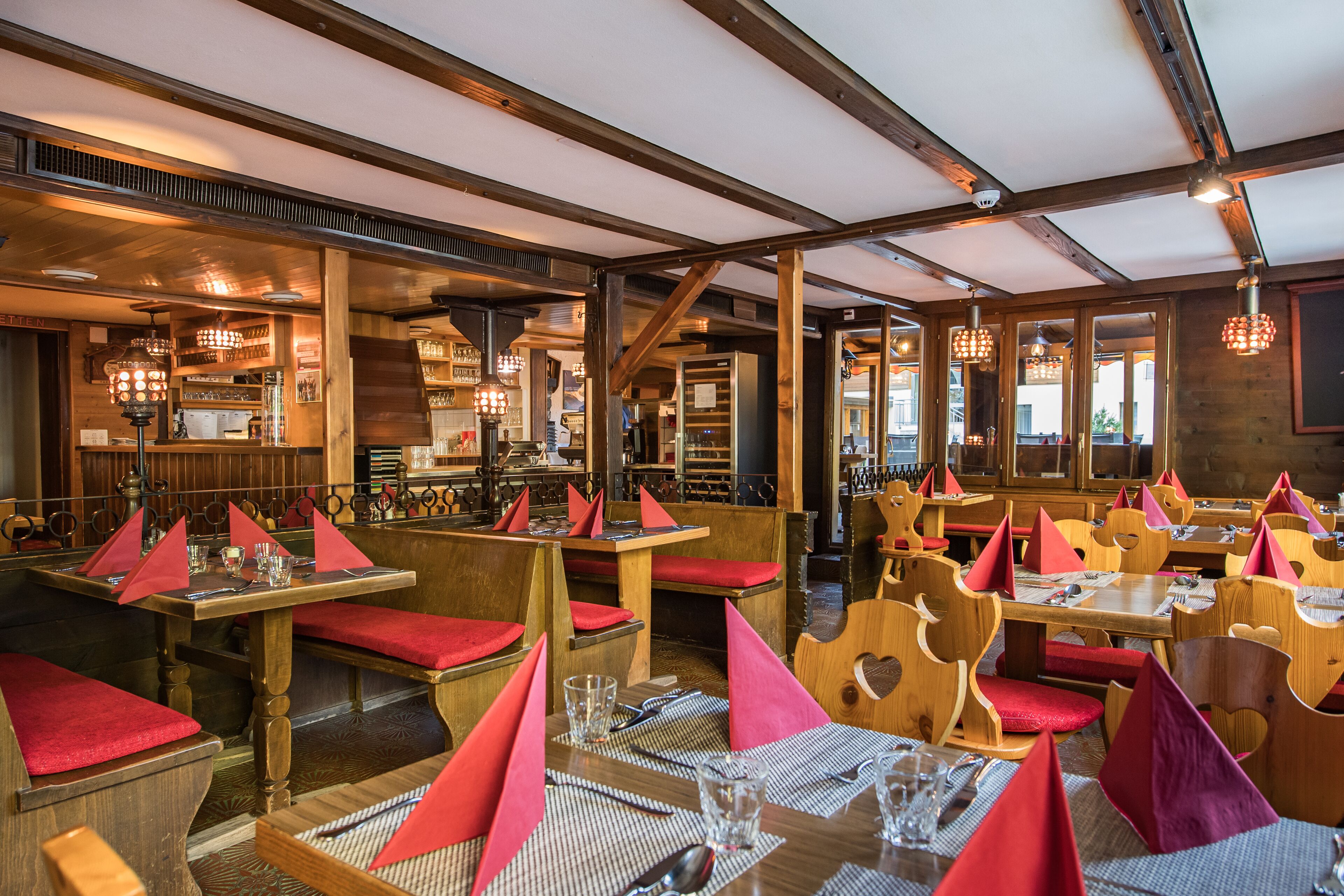 Restaurante com mesas de madeira e bancos estofados vermelhos, apresentando teto alto com vigas aparentes e iluminação pendente.