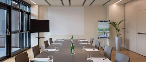 Uma sala de conferências com uma mesa comprida, cadeiras, uma tela de projeção, uma televisão e um vaso de planta.