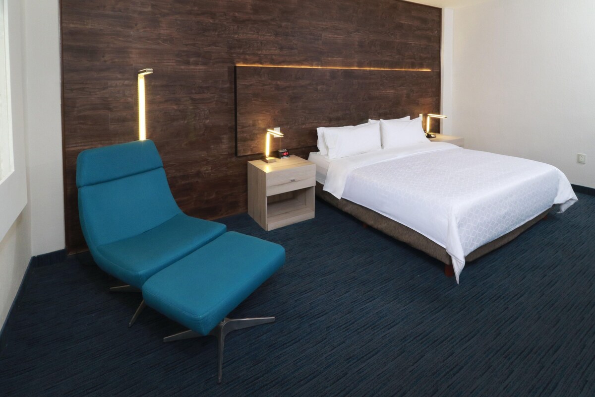 Quarto de hotel com cama, uma cadeira azul, mesa de cabeceira e luminária fixada na parede.