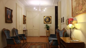 Quarto com escrivaninha de madeira, duas poltronas azuis, tapete estampado, lustre e um quadro na parede.