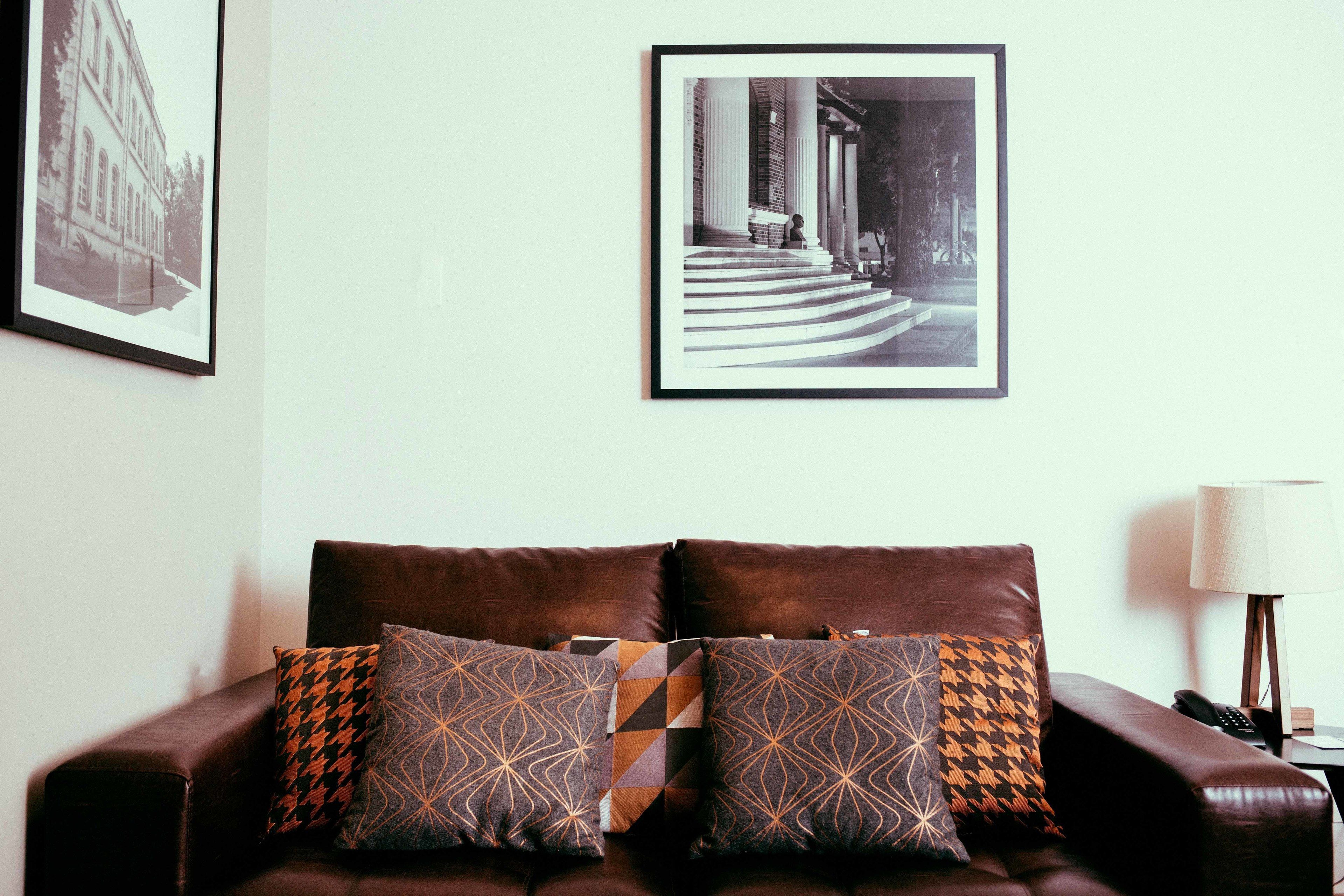 Quarto com sofá de couro marrom, duas almofadas decorativas estampadas, uma fotografia emoldurada na parede e um abajur sobre uma mesinha de cabeceira.