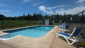 Piscina com espreguiçadeiras, um pequeno armazém e uma área cercada com árvores ao fundo.