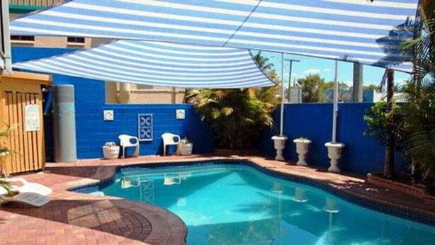 Uma piscina com toldo azul e branco listrado, cercada por um terraço de tijolos e cadeiras de lounge brancas.