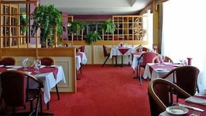 Restaurante com mesas preparadas para refeições, tapete vermelho e grandes janelas com plantas.