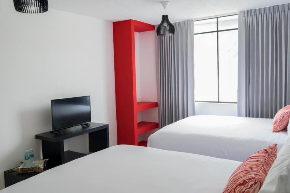 Quarto de hotel com duas camas, uma TV sobre suporte, um aparador vermelho e uma janela com cortinas.