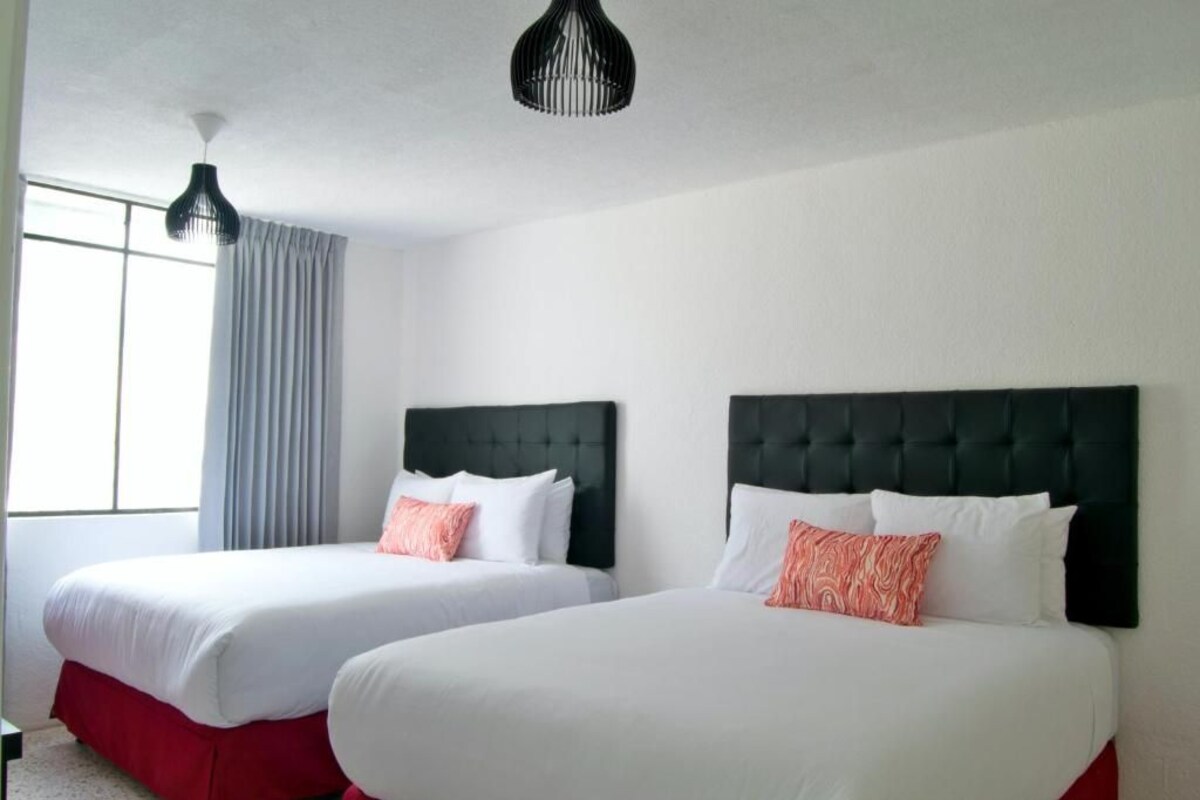Quarto de hotel com duas camas, uma janela grande com cortinas e uma luminária pendente preta.