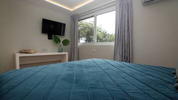 Quarto com cama, televisão na parede, janela com cortinas e uma mesinha com um vaso.