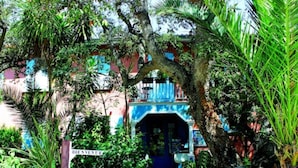 Uma passagem leva a um edifício com fachada azul e vermelha, rodeado por vegetação exuberante e palmeiras.