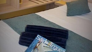 Quarto de hotel com cama, mesa de cabeceira, abajur, cabideiro e uma revista sobre a cama.
