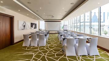 Uma sala de conferência com carpete verde, cadeiras brancas, mesas, uma tela de projetor e um quadro branco.