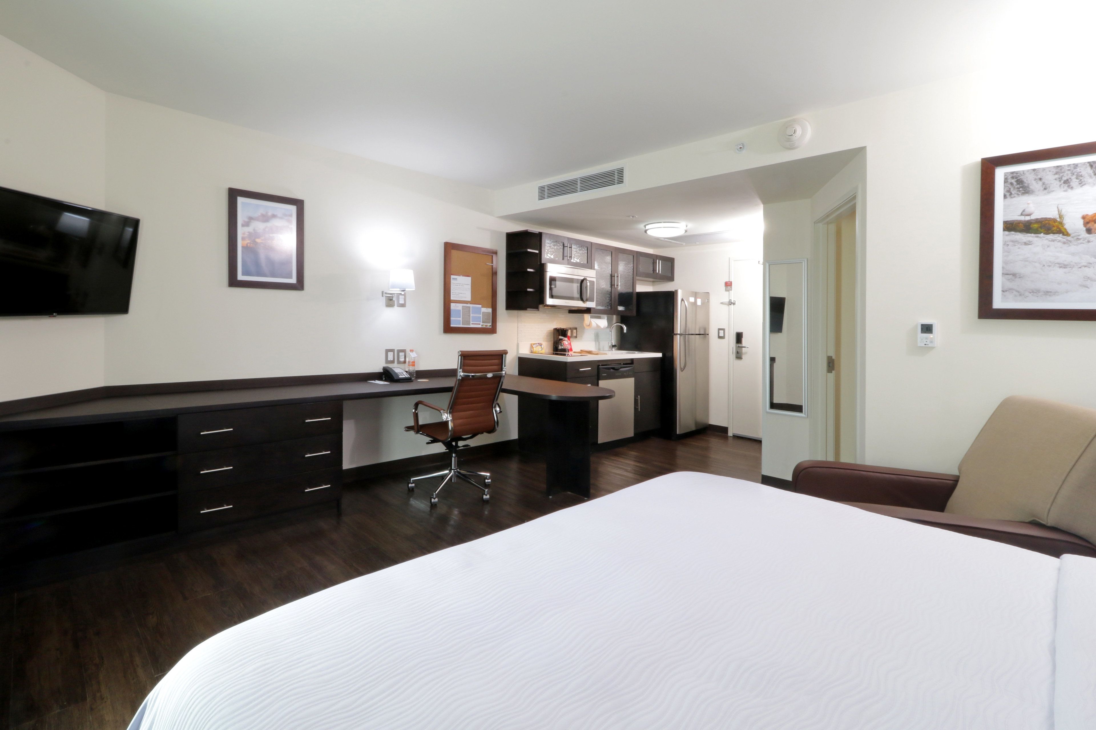 Quarto de hotel com uma cama grande, uma escrivaninha com cadeira, cozinha compacta e televisão na parede.