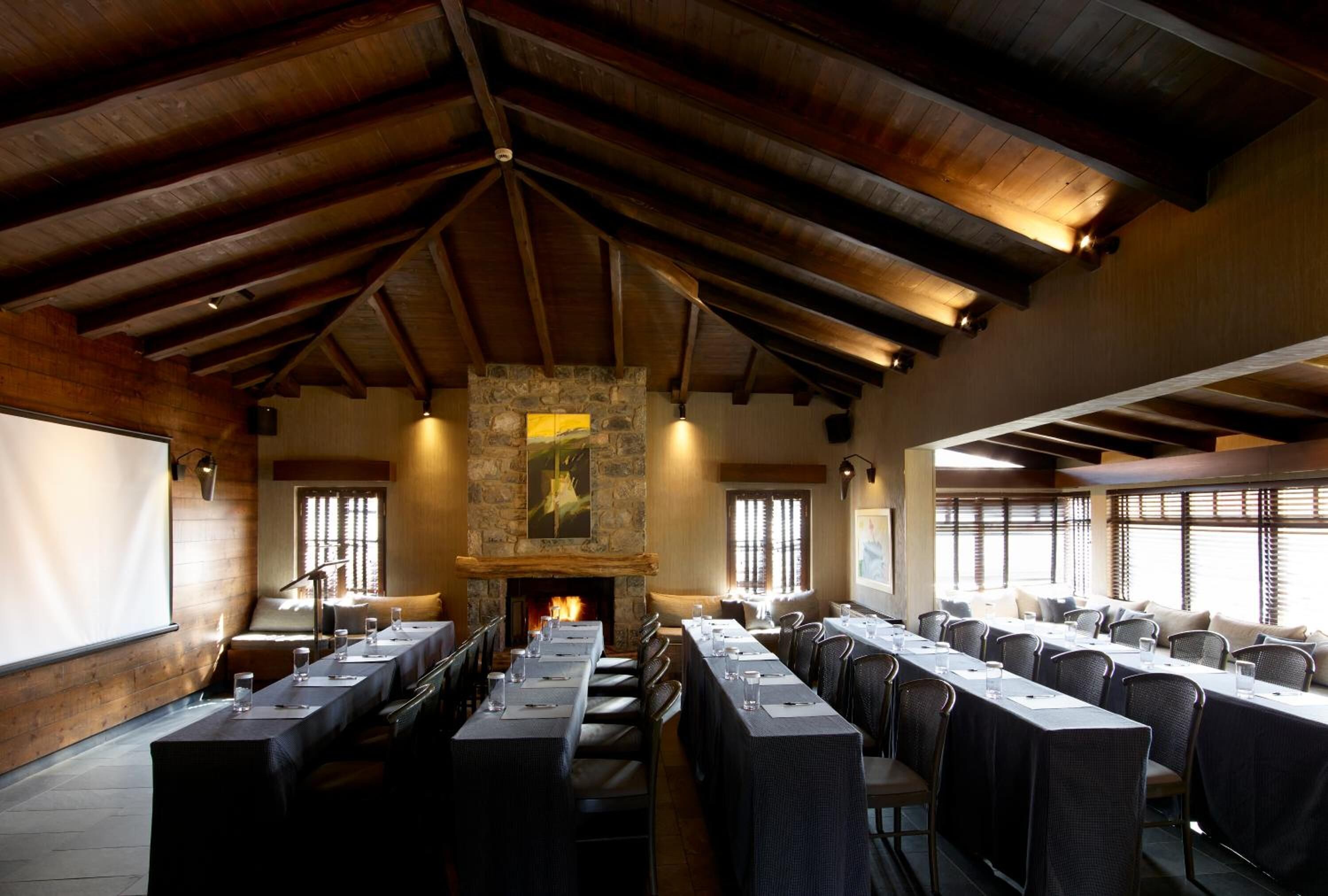 Uma sala de conferências com mesas longas, cadeiras, uma lareira de pedra e vigas de madeira.