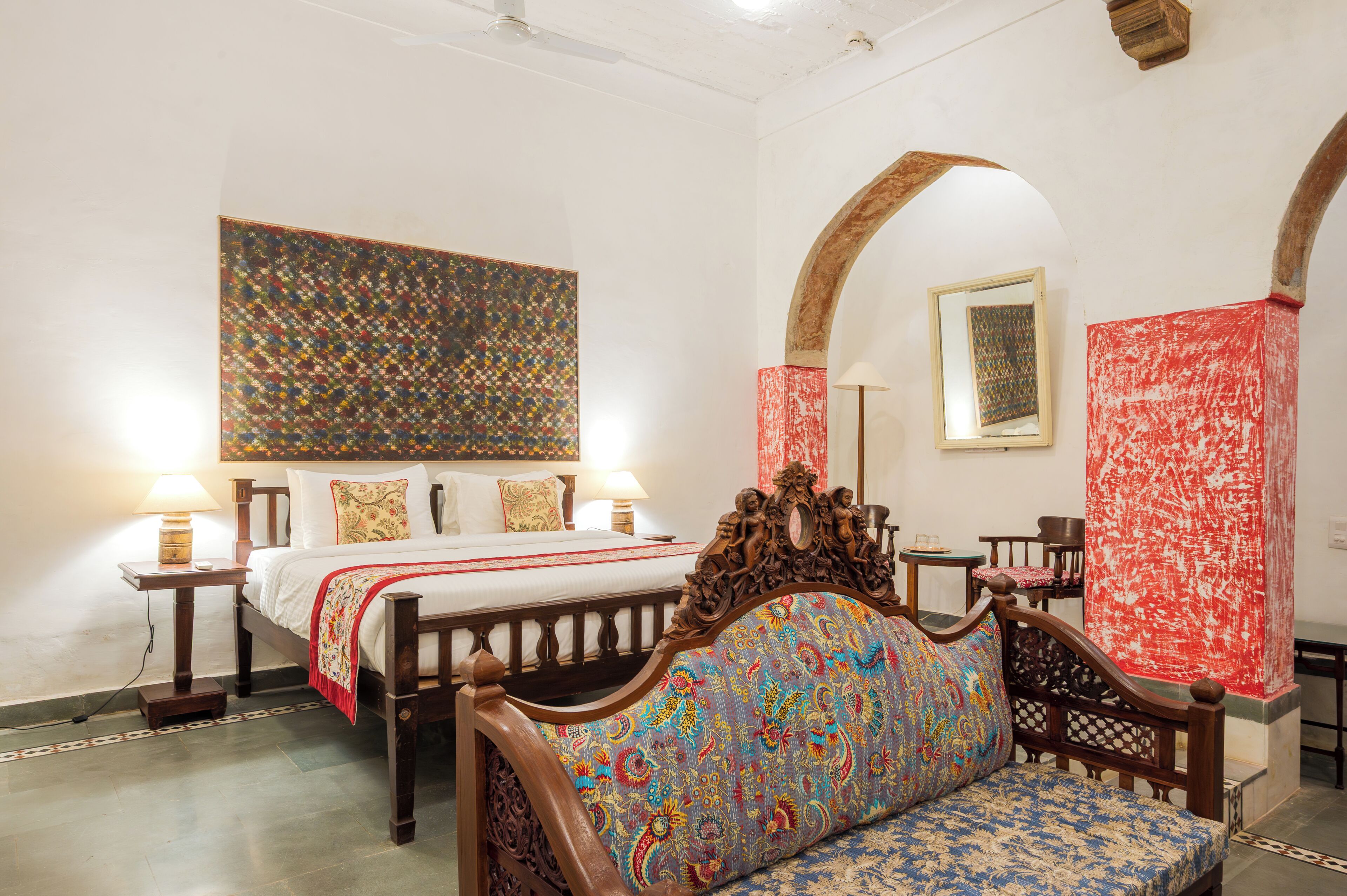 Quarto com cama grande, cabeceira de madeira, sofá estampado e cortina vermelha.