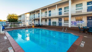 Piscina com borda de tijolos, um edifício branco com varandas e uma placa indicando que mergulhos não são permitidos.