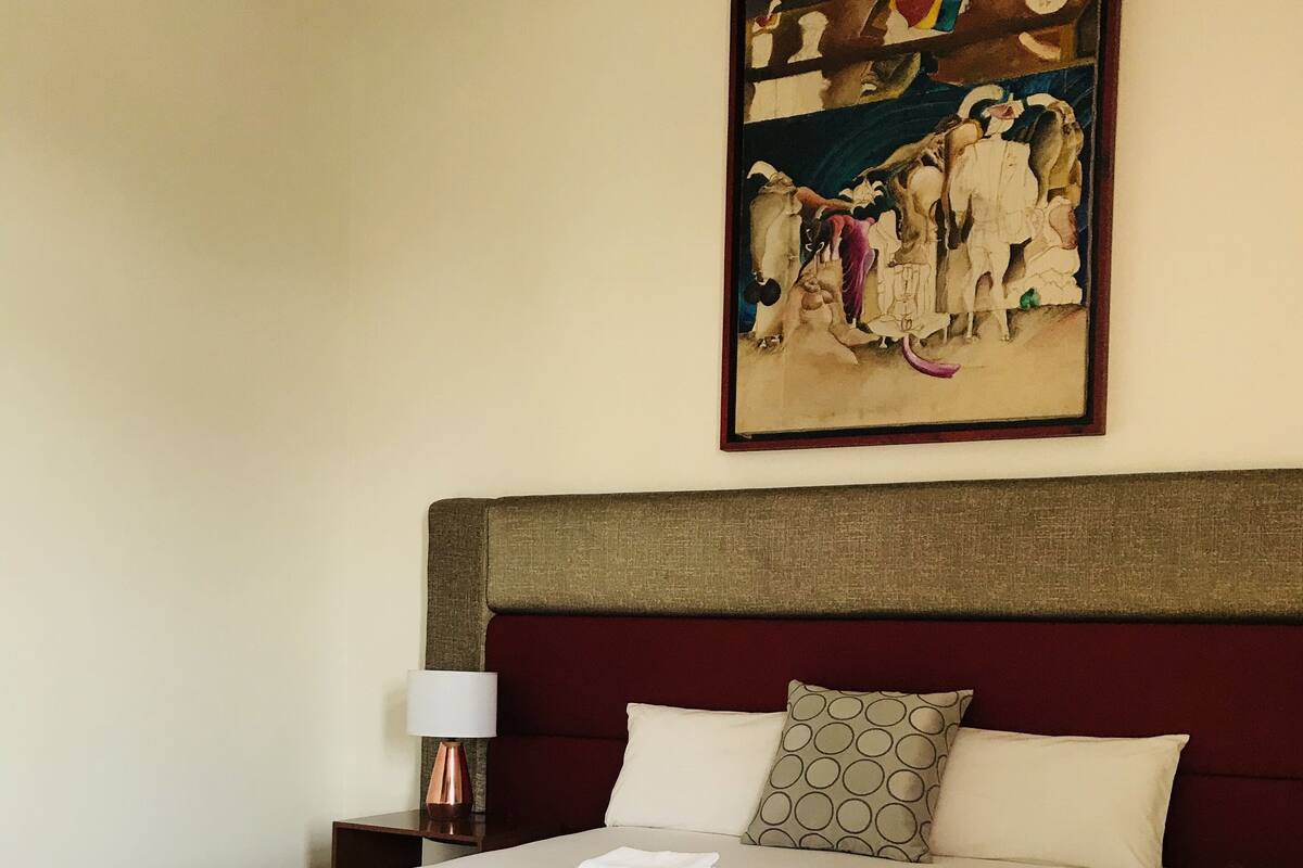 Quarto de hotel com uma cama grande, cabeceira, mesa de cabeceira, abajur e um quadro na parede.